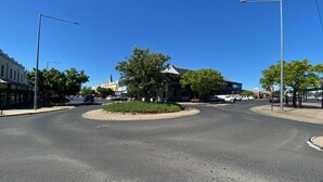 Uma rotatória com área ajardinada central, rodeada por edifícios com estabelecimentos comerciais à esquerda e um edifício moderno e amplo à direita.