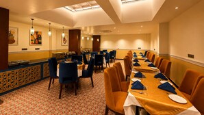 Uma sala de jantar de hotel com uma mesa comprida posta para refeição, várias cadeiras e uma área de buffet com equipamentos de serviço de alimentos.