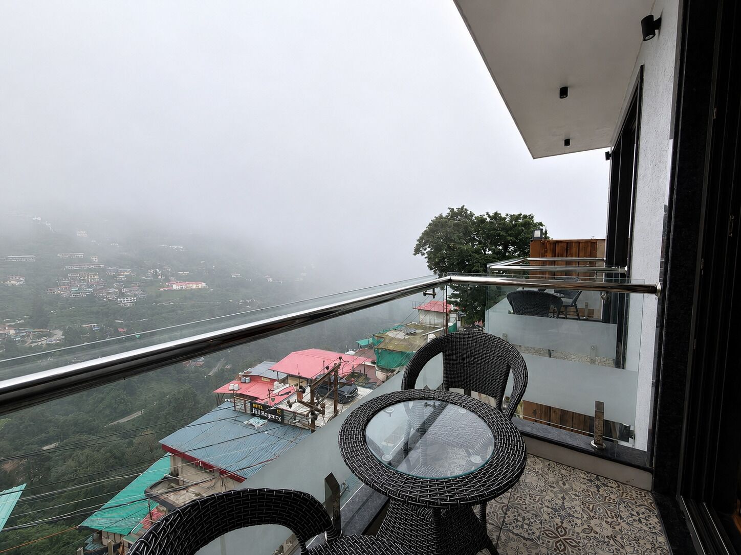 Uma varanda com mesa e cadeiras de vidro, com vista para prédios e uma árvore.