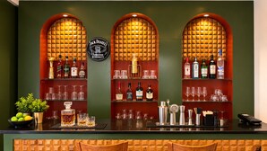 Área de bar com parede verde, três nichos em arco exibindo garrafas de bebidas e dois banquinhos de madeira.