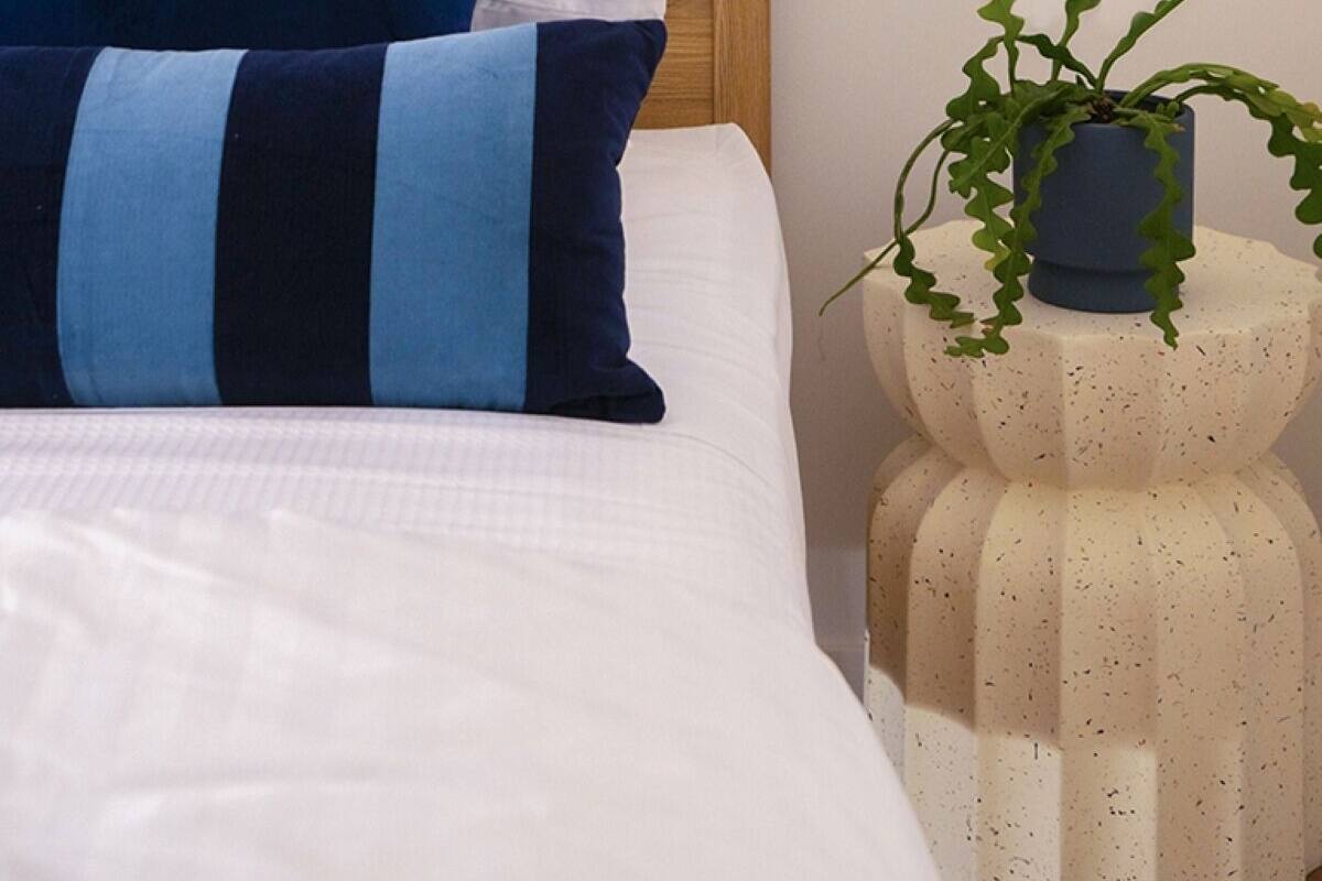 Uma cama bem arrumada com cabeceira de madeira, um abajur e um pequeno vaso com planta em uma mesa redonda ao lado da cama.