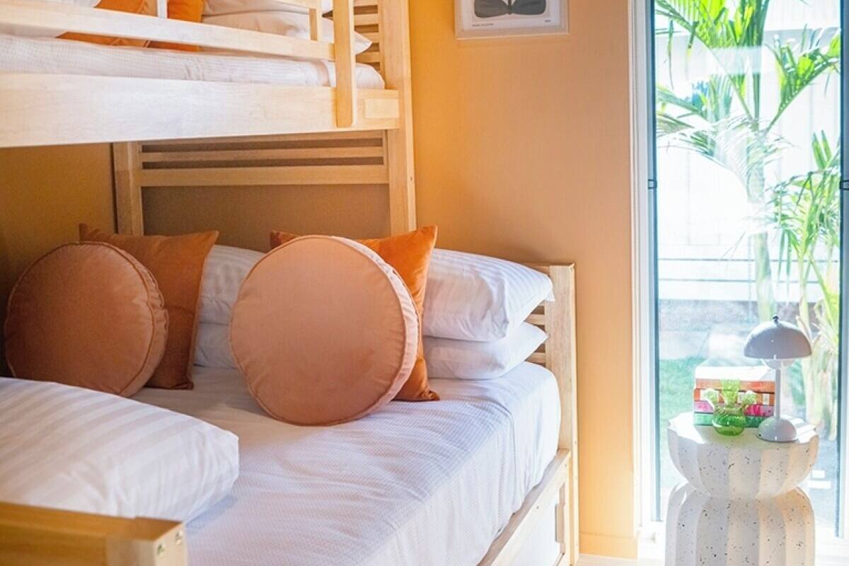 Uma cama beliche com roupa de cama branca, uma mesinha com abajur, e uma janela com vista para o verde.
