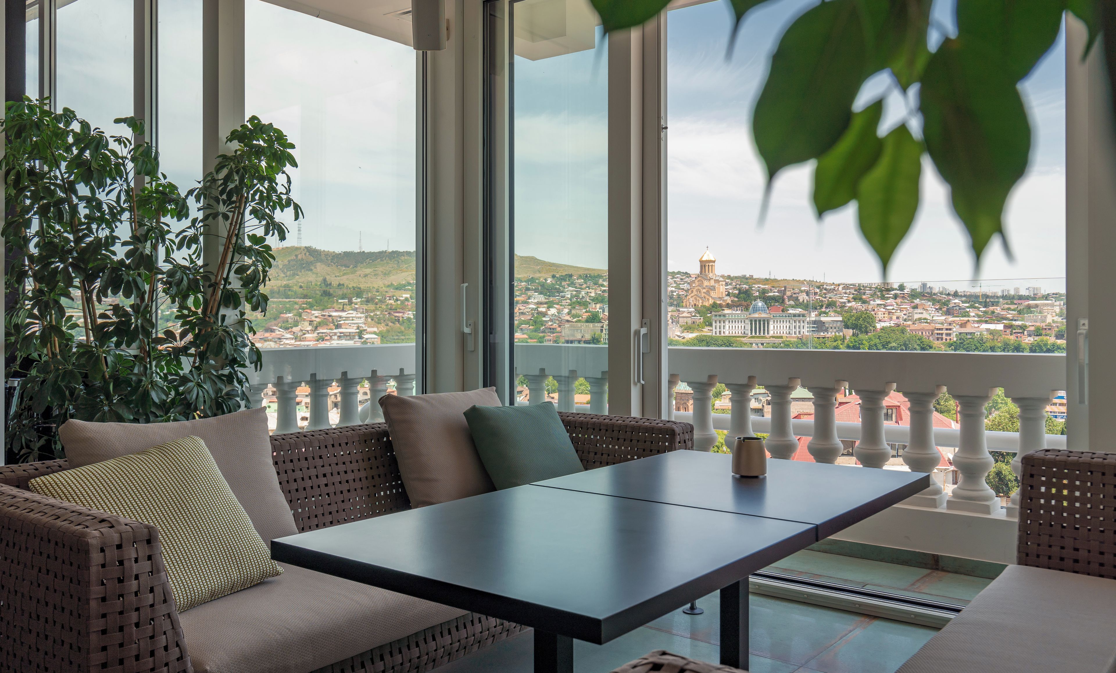 Varanda com sofá e mesa, vista para um horizonte urbano com edifícios e uma colina.