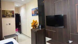 Quarto de hotel com uma cama, escrivaninha, televisão, planta e um quadro na parede.