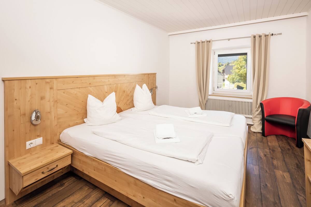 Quarto com cabeceira de madeira, duas camas, uma cadeira vermelha, janela com cortinas e piso de madeira.