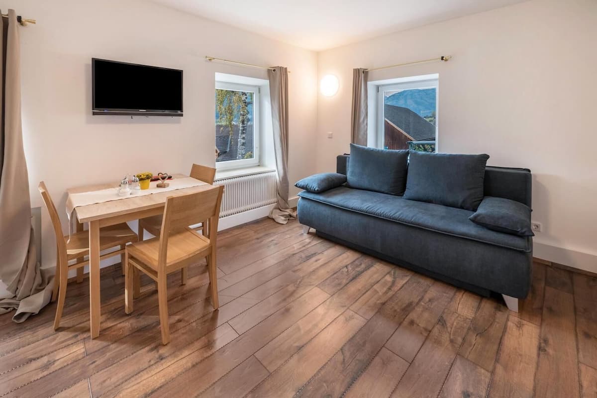 Quarto com mesa de jantar de madeira, um sofá, televisão na parede e duas janelas com cortinas.
