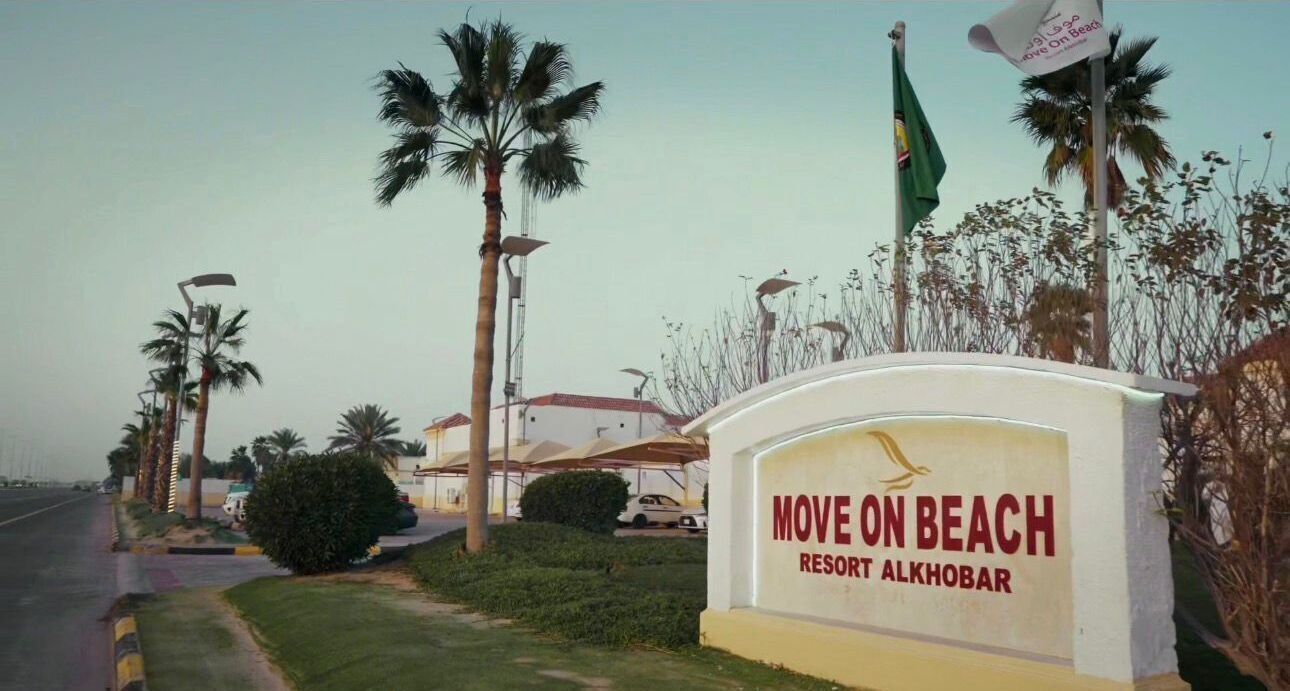 Placa para 'MOVE ON BEACH RESORT ALKHOBAR' com palmeiras e um edifício ao fundo.