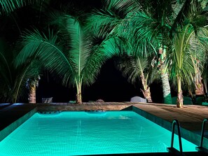 Piscina iluminada à noite com um deque de madeira e palmeiras.