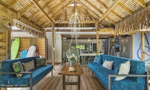 Interior de madeira com teto alto, dois sofás estampados em azul, uma mesa de madeira e uma prancha de surf encostada na parede.