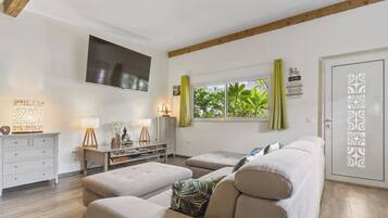 Uma sala de estar com sofá, pufes, televisão, janela com cortinas e um armário.