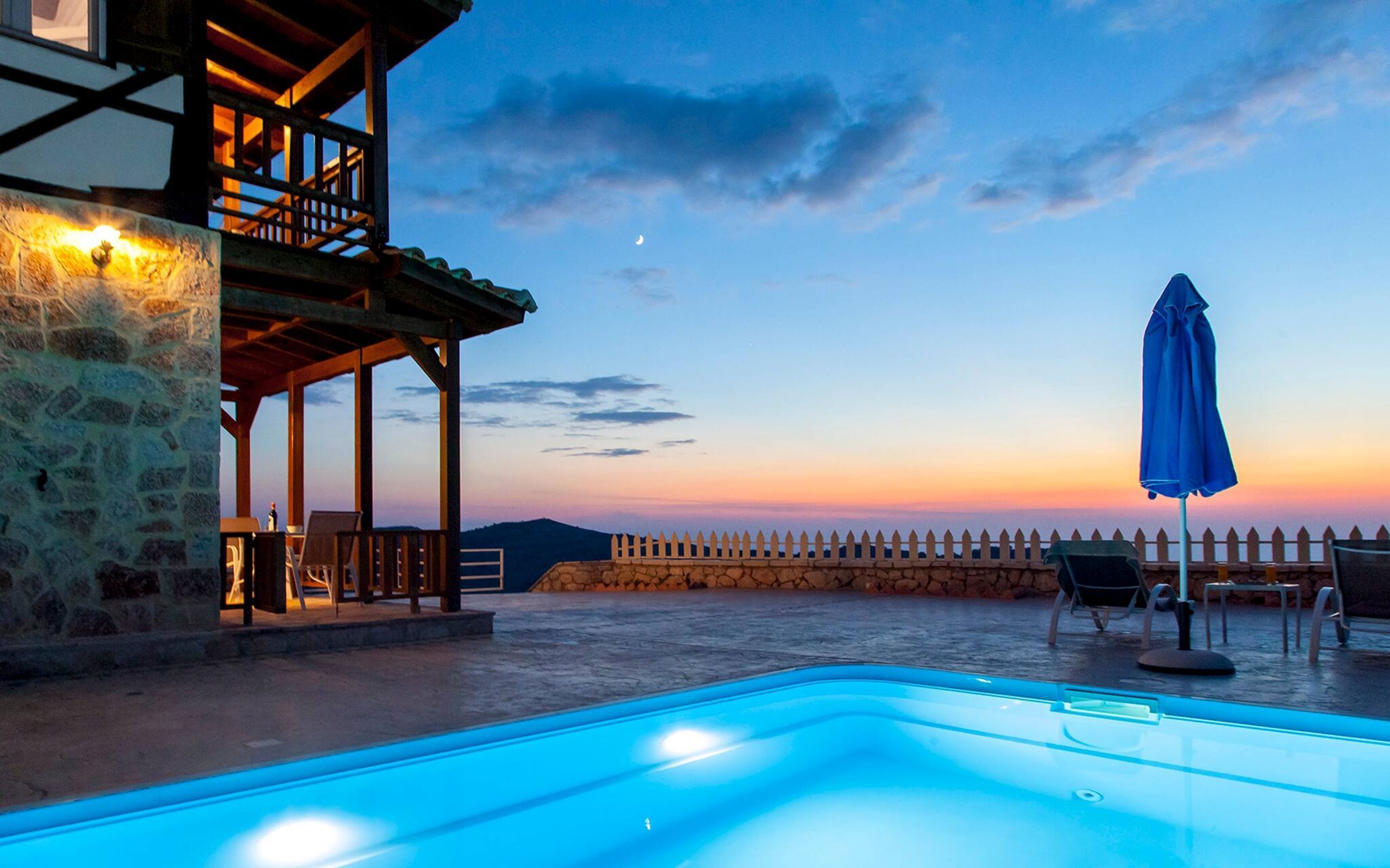 Área da piscina com muro de pedra, deck de madeira e vista para o mar ao pôr do sol.