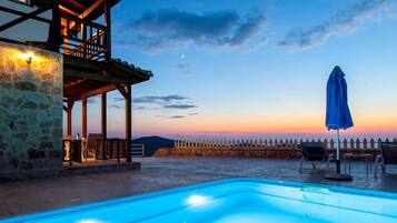 Área da piscina com muro de pedra, deck de madeira e vista para o mar ao pôr do sol.