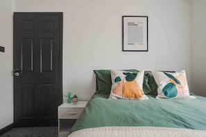 Quarto com porta escura, uma obra de arte emoldurada na parede, cama com roupa de cama verde e uma pequena planta no criado-mudo.