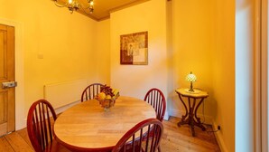 Uma sala de jantar com mesa de madeira, cadeiras, uma tigela de frutas, um candelabro, um quadro e um abajur.