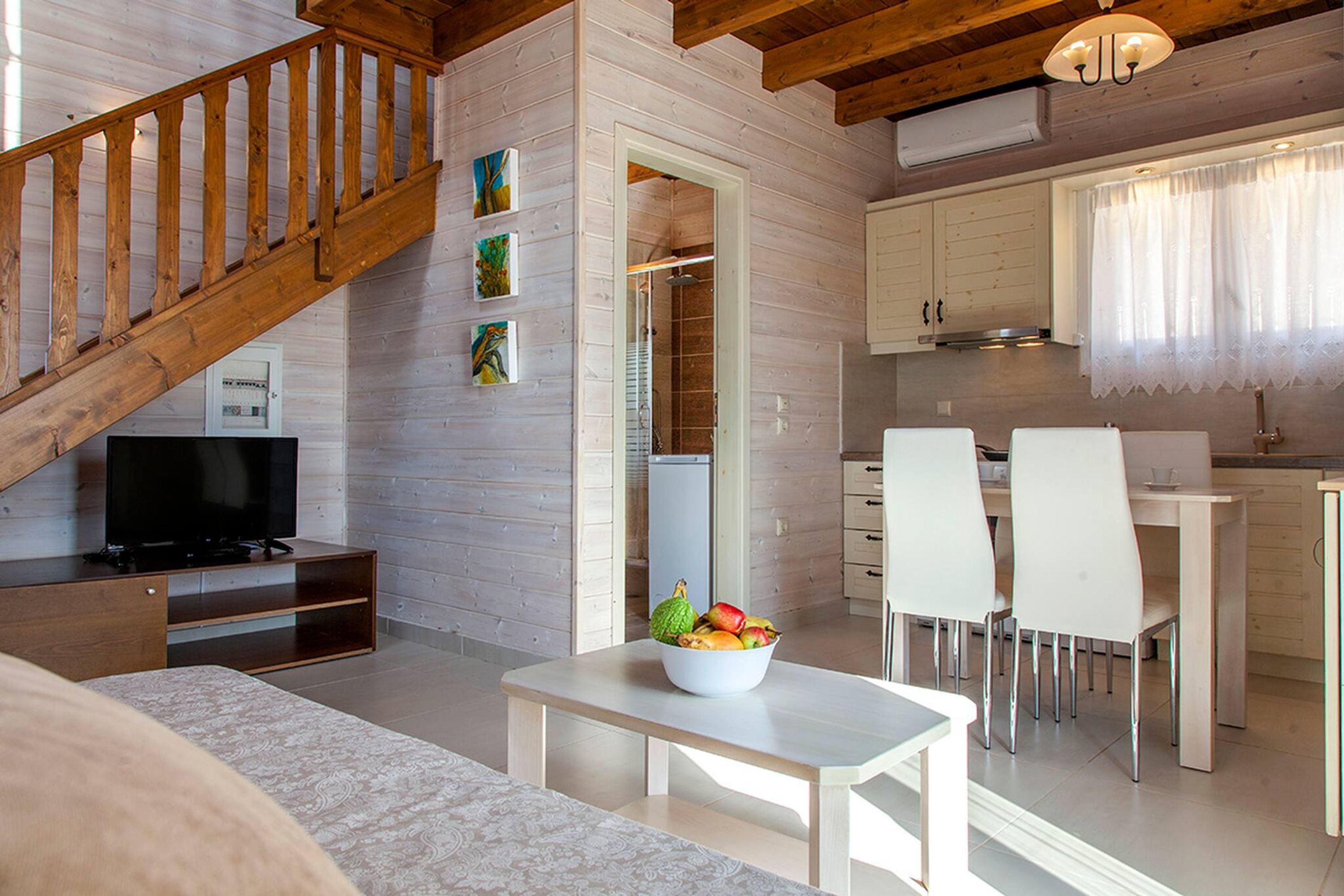 Um interior moderno com escada de madeira, área de jantar com cadeiras brancas, cozinha com armários brancos e uma televisão sobre um suporte de madeira.