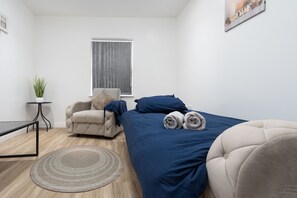 Quarto com cama, uma cadeira, uma mesinha, um vaso de planta e uma janela com cortinas.