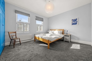 Quarto com cama, cadeira, mesa de cabeceira, luminária, radiador e janela com persianas.
