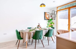 Uma área de refeições com uma mesa redonda, cadeiras verdes, um banco de madeira e uma janela grande.