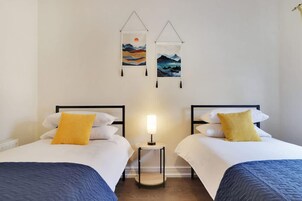 Quarto de hotel com duas camas, cada uma com uma almofada amarela, uma pequena mesa de cabeceira com abajur e obras de arte nas paredes acima das camas.