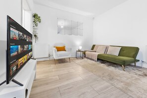 Uma sala de estar moderna com uma TV de tela plana, uma poltrona branca, um sofá verde e uma mesinha lateral.