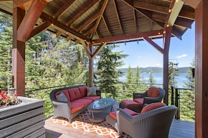 Um deck coberto ao ar livre com móveis de vime, uma mesa de vidro e vista para um lago e floresta.
