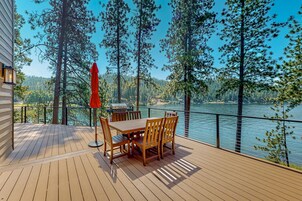Um deck de madeira com mesa e cadeiras, com vista para um lago, árvores e uma casa ao longe.