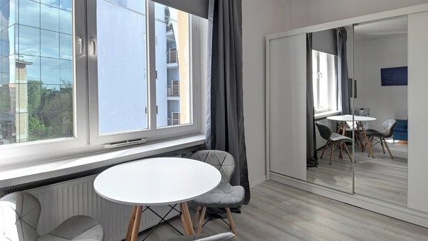 Quarto moderno com uma mesa redonda, duas cadeiras cinzas, uma janela ampla e uma porta de vidro deslizante para outro espaço.