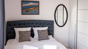Cama bem arrumada com lençóis brancos e duas almofadas, um espelho redondo na parede e um quadro acima da cama.