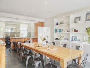 Uma cozinha moderna com uma mesa de jantar de madeira, banquetas de bar e uma ilha central. Há estantes com livros e objetos decorativos.