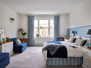 Quarto com cama, sofá azul, penteadeira, mesa de cabeceira, janela com cortinas e vista para o exterior.