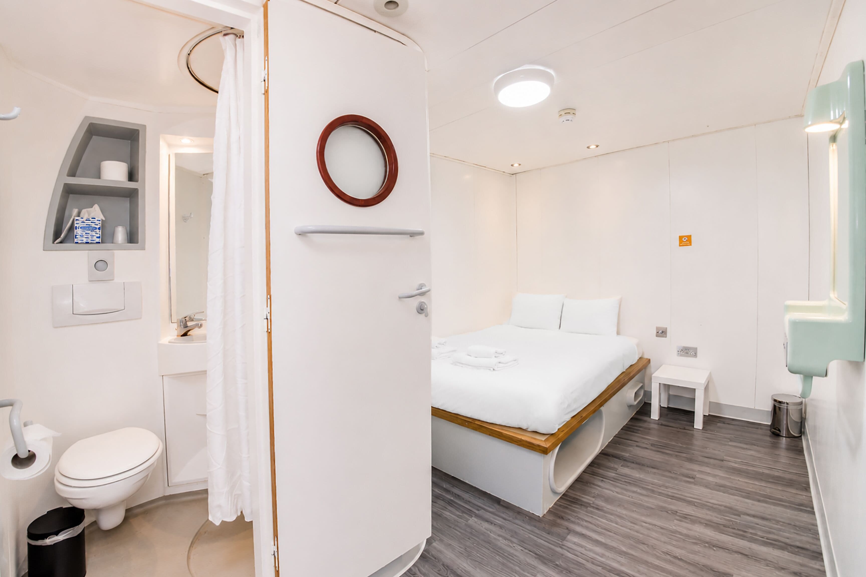 Quarto de hotel com casa de banho, incluindo sanita, lavatório, chuveiro, cama, mesa de cabeceira e um pequeno banquinho.