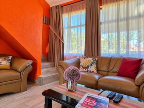 Uma sala de estar com sofá, mesa de centro com revista e controles remotos, escada e janela com cortinas.