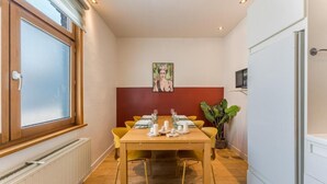 Uma sala de jantar com mesa de madeira posta para quatro lugares, cadeiras amarelas, um vaso com planta e um quadro na parede.