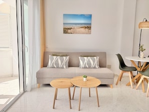 Uma sala de estar moderna com um sofá, duas mesas redondas de madeira, uma mesa de jantar com cadeiras, uma fotografia de uma praia e uma janela com cortinas.
