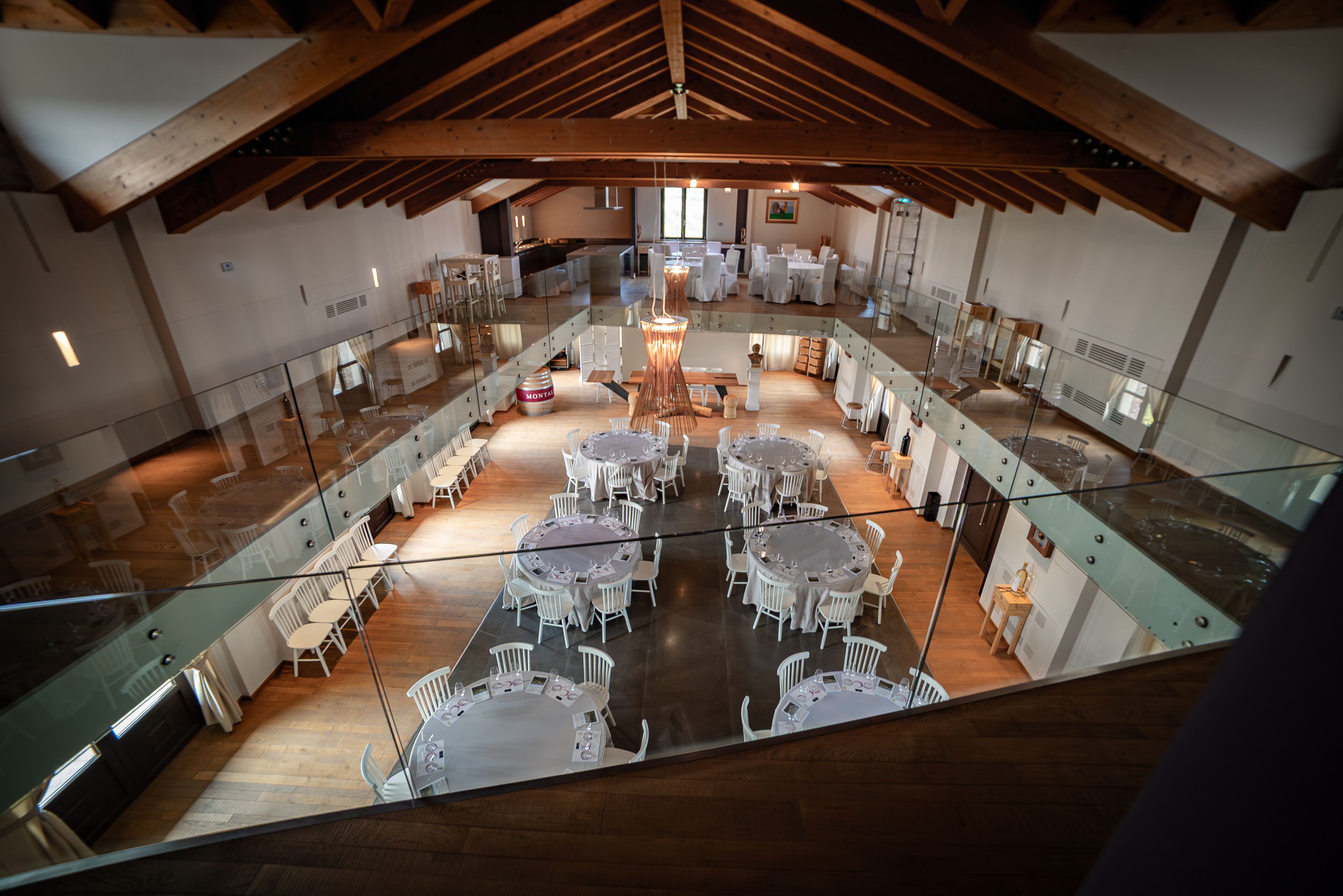 Uma sala de jantar espaçosa com várias mesas e cadeiras redondas, teto alto com vigas de madeira e vista para a cozinha.