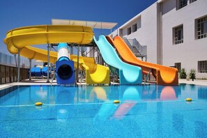 Piscina com várias escorregas aquáticas em cores variadas, céu azul límpido e um edifício com varandas ao fundo.
