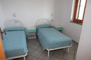 Quarto pequeno e limpo com duas camas de solteiro, cada uma com cabeceira e peseira combinando, e uma janela com moldura de madeira.