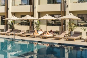 Área de piscina com espreguiçadeiras e guarda-sóis, piscina cristalina e fachada moderna de um edifício.