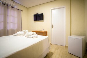 Quarto de hotel com cama, uma pequena mesa, televisão fixada na parede, porta e mini-geladeira.