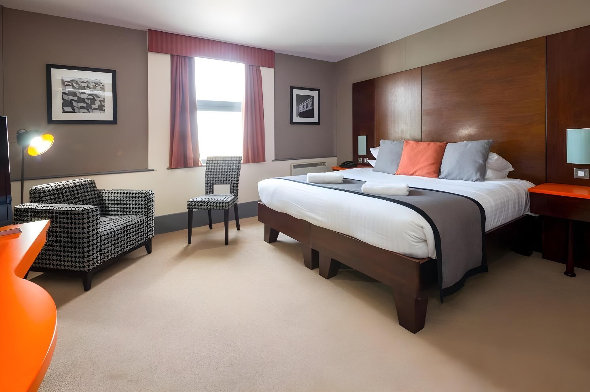 Quarto de hotel com cama, uma cadeira, escrivaninha, abajur e duas quadros na parede.