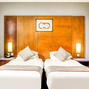 Quarto de hotel com duas camas, cabeceira de madeira, quadro emoldurado, abajures e telefone na mesinha de cabeceira.