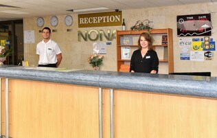 Um balcão de recepção com uma placa que diz 'RECEPTION NONA', um relógio e um visor digital mostrando a hora e a data.