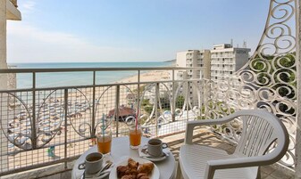 Varanda com mesa posta para dois, com vista para uma praia com guarda-sóis e um edifício com a placa 'ELITSA' ao fundo.