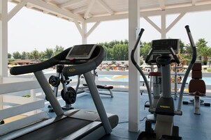 Uma academia coberta ao ar livre com esteiras e equipamentos de exercício, com vista para uma piscina com guarda-sóis e espreguiçadeiras.