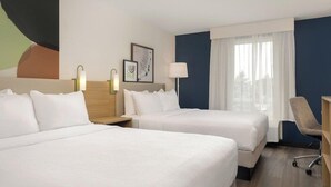 Quarto de hotel com duas camas, uma escrivaninha, uma cadeira e uma janela grande com cortinas.