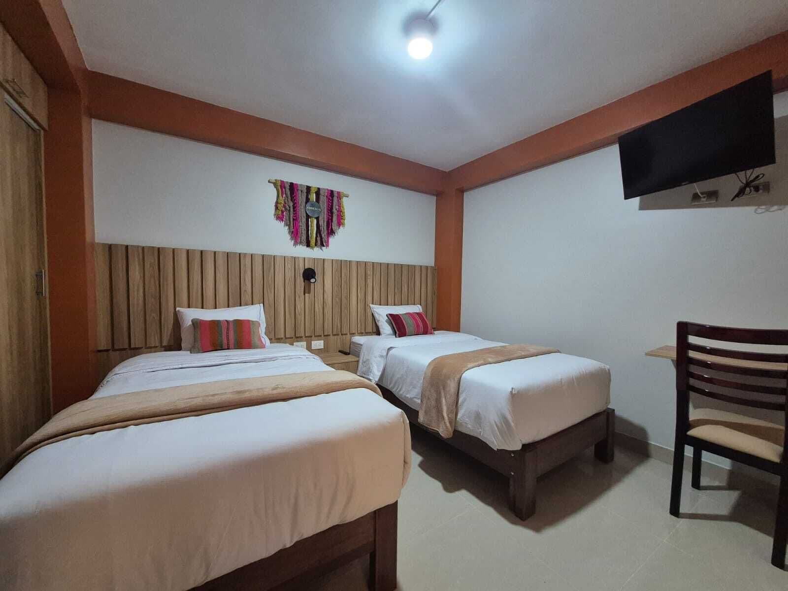 Quarto de hotel com duas camas, cabeceira de madeira, televisão fixada na parede e uma cadeira.