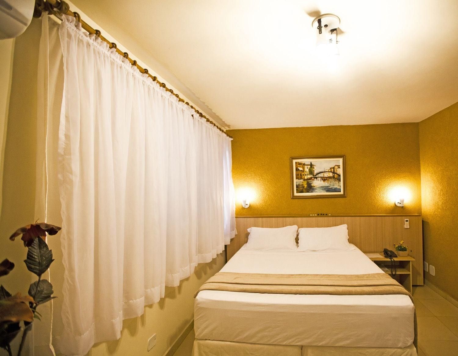 Quarto de hotel com cama, cortinas, um quadro e uma mesa de cabeceira.