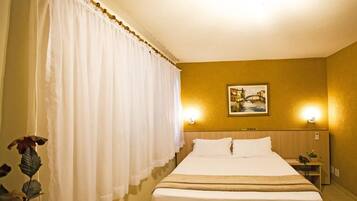 Quarto de hotel com cama, cortinas, um quadro e uma mesa de cabeceira.