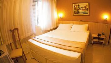 Quarto de hotel com uma cama grande, uma cadeira, mesa de cabeceira com abajur, um quadro na parede e uma janela com cortinas.
