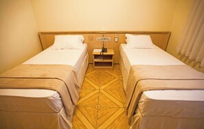 Quarto de hotel com duas camas de solteiro, piso de madeira, mesa de cabeceira com telefone e interruptor de luz fixado na parede.